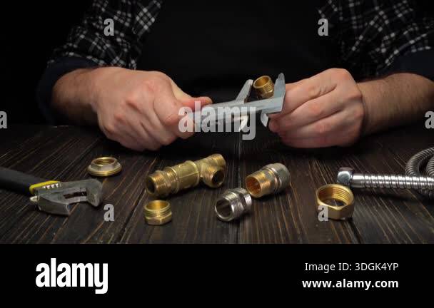 A plumber holds a caliper and measures brass fittings on a wooden table ...