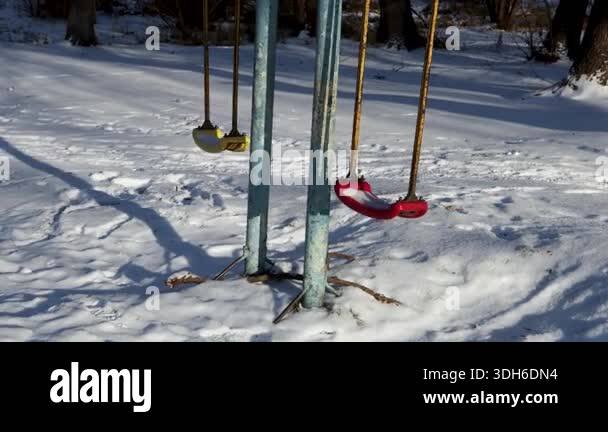 In a quiet snowy park, empty swings move gently on a clear winter day ...