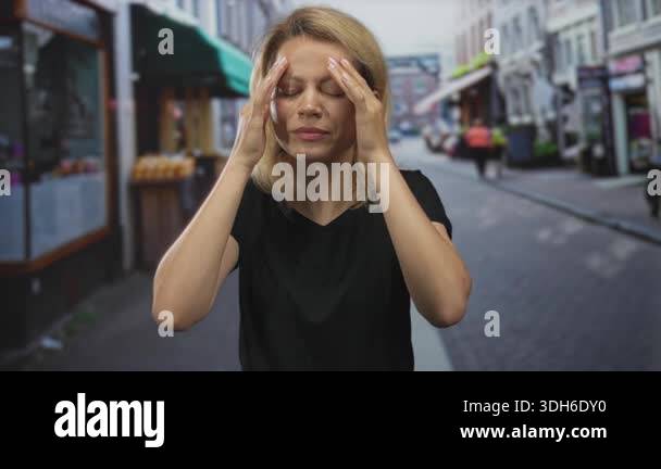 Young blonde woman pressing hands to forehead on busy city street with ...