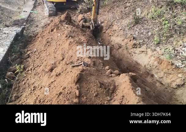 An excavator uses its bucket to dig a narrow trench for laying pipes ...