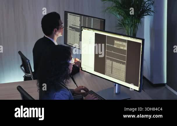 Professionals analyzing coding data on dual monitors in a dim modern ...