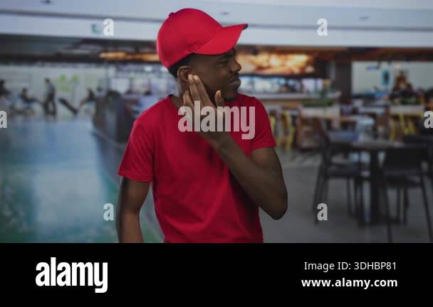 Young courier in red shirt and cap touches ear and winces while ...