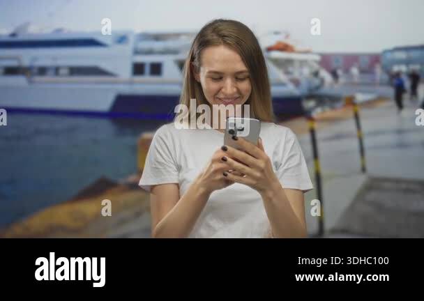 Woman smiling at smartphone while standing near boat at port, capturing ...
