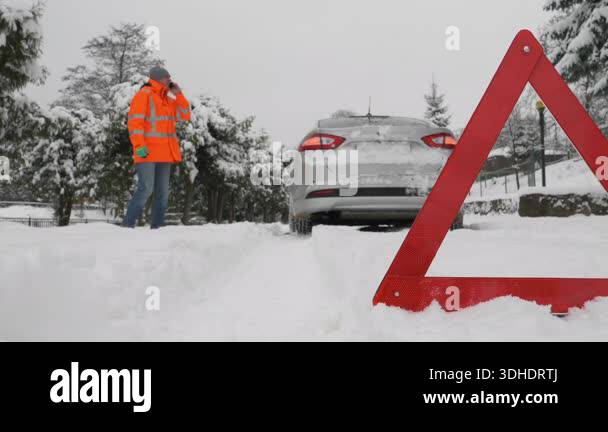 A person in an orange jacket stands near a car stuck in deep snow. A ...