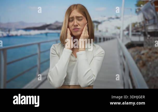 Young blonde woman holding cheek with both hands on sunlit seaside ...