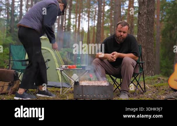 Young man and woman enjoying a relaxing picnic in the woods, grilling meat on a barbecue while ...
