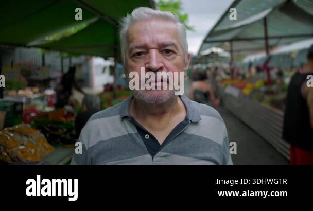 Older man pauses between produce stalls and looks directly at camera ...