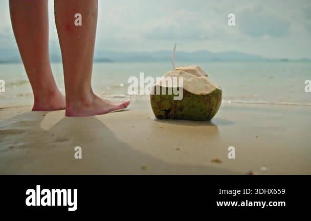 Barefoot person standing on sandy beach beside green coconut with straw ...