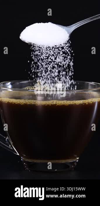 A spoonful of sugar is poured into a glass cup of tea against a dark ...