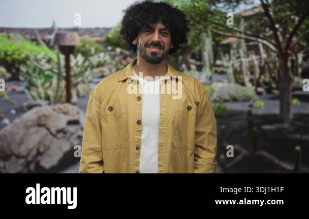 Man with curly hair and beard salutes while smiling, wearing yellow ...