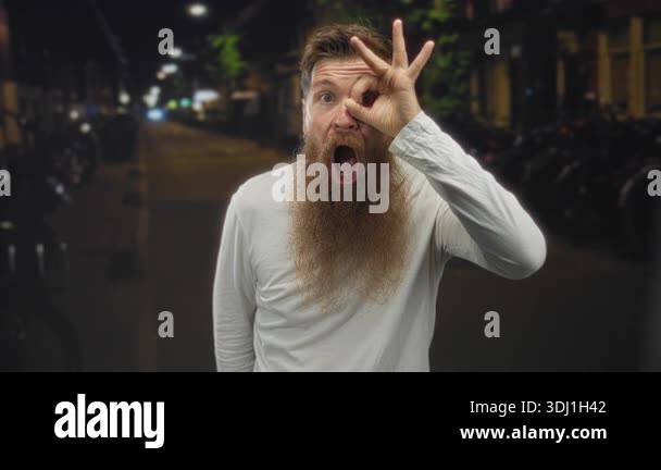 Man with long bushy beard makes ok sign with hand framing eye on dimly ...