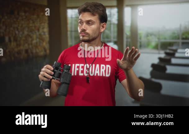 Man lifeguard holding binoculars and whistle scanning an indoor pool ...