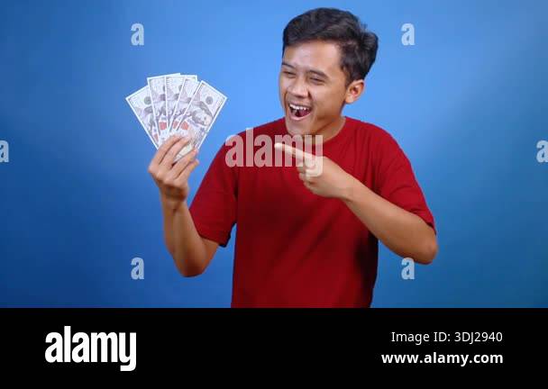 Cheerful young man pointing at a fan of hundred-dollar bills against a ...