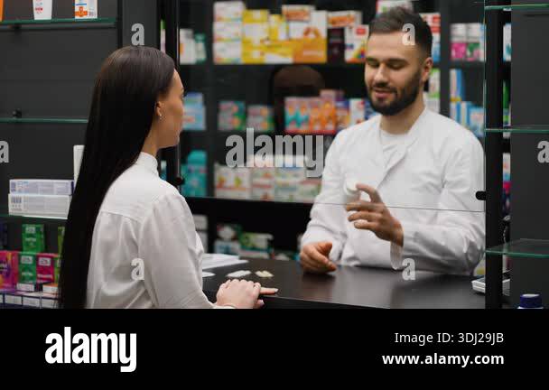 Young male pharmacist standing behind the counter, talking to a female ...