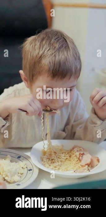 Preschool child eats noodles with gusto, watched by a small pet in a ...