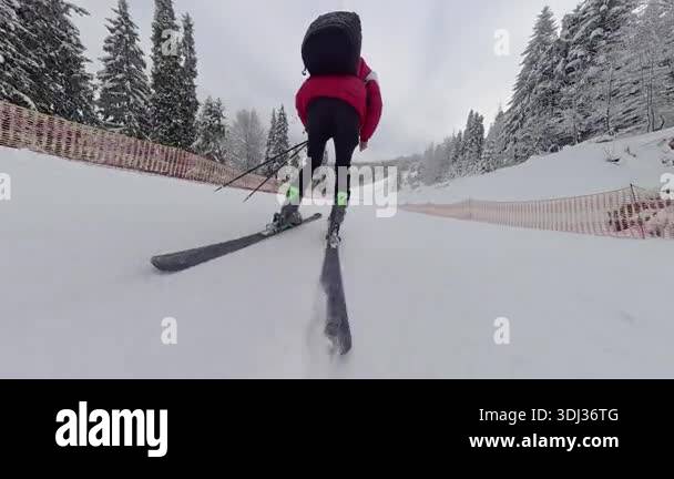 Rear view of a skier in a red jacket riding down a groomed snowy slope ...