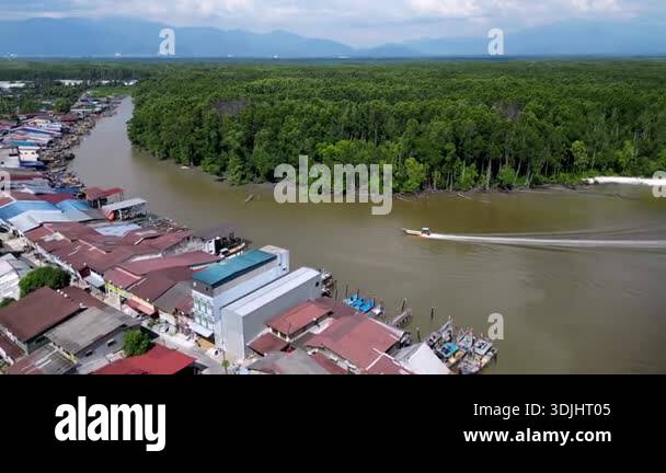 A boat is traveling down a river near a town. The river is brown and ...