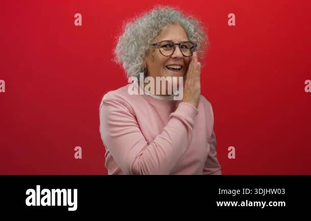 Woman whispering in a playful manner on a solid red background wearing ...