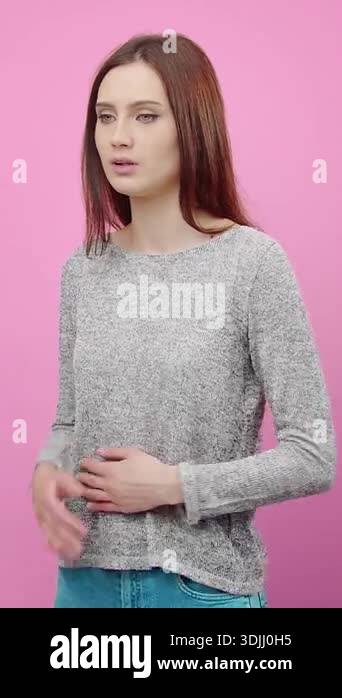 Woman in gray shirt holds her stomach against pink background, showing ...
