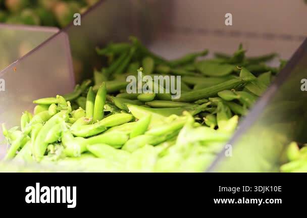 A hand picks green peas from a supermarket bin under bright lighting ...