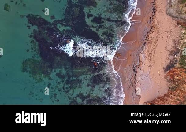 Overhead view of a sandy beach with waves crashing on rocks. Clear blue ...