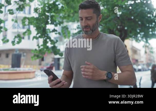 Man touching neck and holding smartphone on a tree lined city street ...