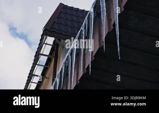 Icicles hang from the edge of a dark roof against a cloudy sky. Low ...