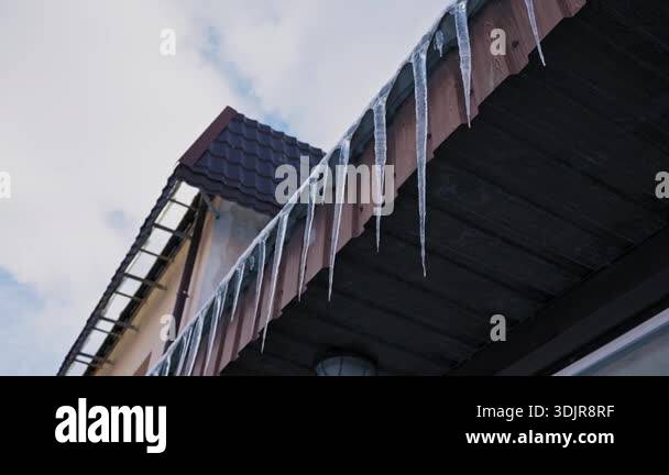 Long, sharp icicles hang in a row from the eaves of a house roof. White ...