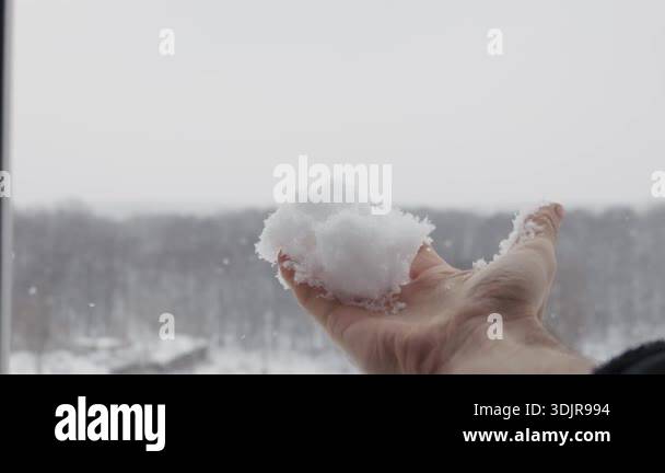 A persons hand in squeezes a clump of fresh snow in front of a window ...