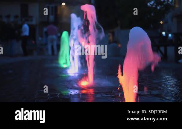 Illuminated city fountain with tall water jets against architecture of ...