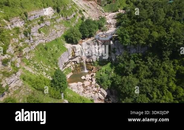Aerial drone follows a mountain stream over a cliff to a waterfall by a ...