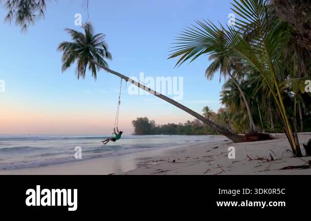 Gentle waves lap at the shore as a woman swings from a palm tree in Koh ...