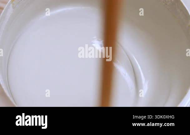 A close-up of a stirring stick stirring shiny white wall paint in a ...