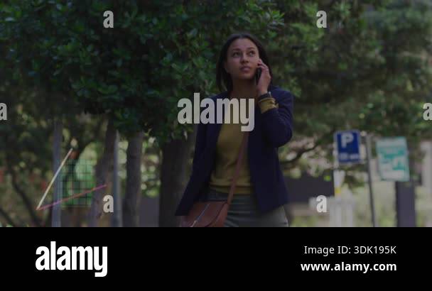 Businesswoman walking leafy sidewalk talking on smartphone while ...