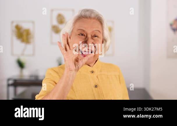 Asian grandmother in yellow shirt smiles and makes an OK hand gesture ...
