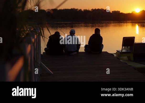 Group of friends fishing on wooden pier during golden sunset by calm ...