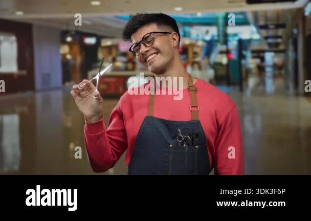 Young hispanic man in apron and glasses holding scissors at a shop in a ...