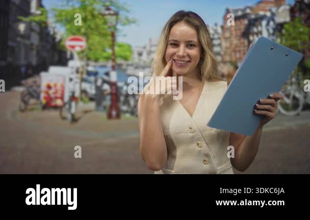 Woman points finger to cheek while holding clipboard on street in ...