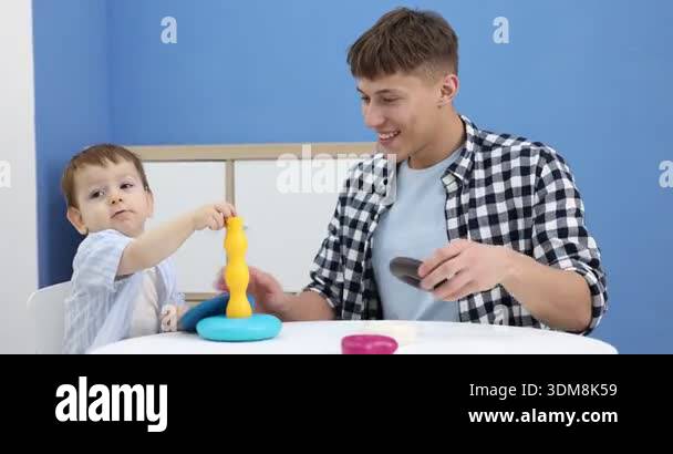 Smiling male nanny and cute little boy playing with toys at white table ...