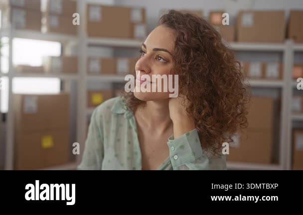 Young hispanic woman with hand on chin beside stacked parcel boxes on ...