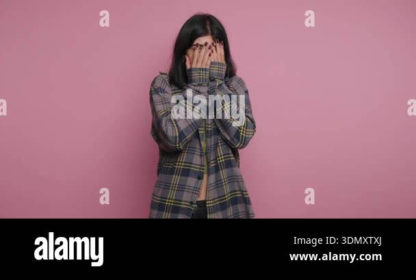 A woman stands in front of a pink wall with her hands covering her face ...
