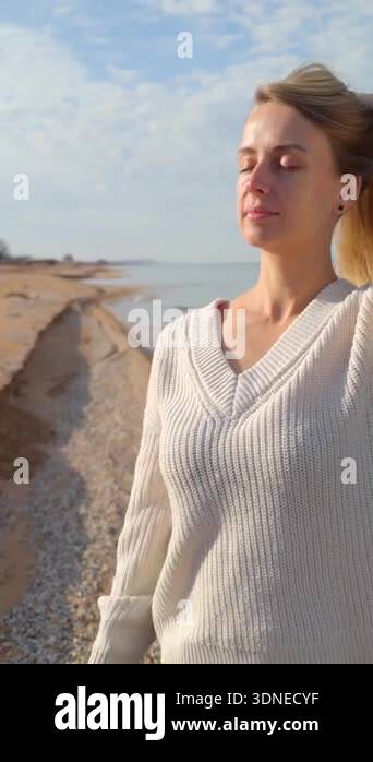 Young woman standing on a sandy beach near the sea, adjusting her hair ...