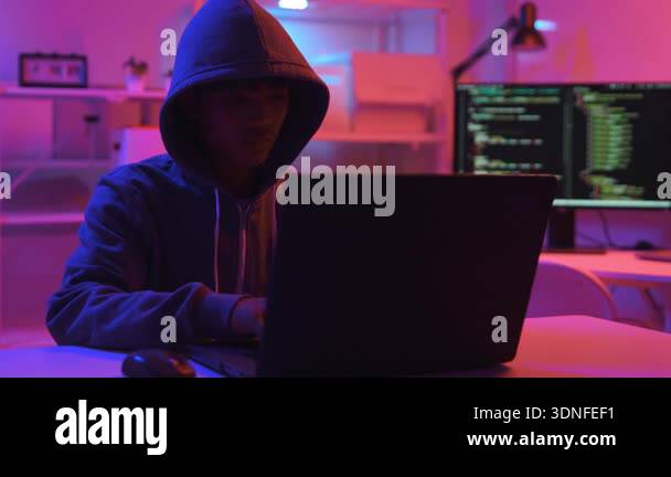 the man in a hood working as a hacker at the computer in the dark room ...