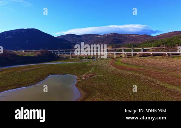 A large bridge extends over a river in a valley. Nearby hills rise in ...