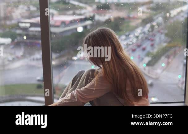 Sad girl sitting at the panoramic window on a cloudy rainy day and ...