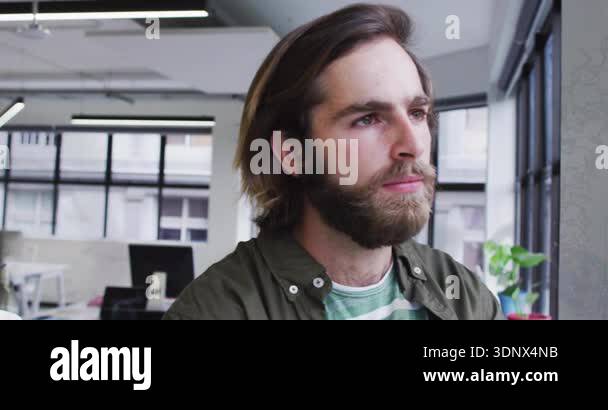 Office worker standing at window, turning head, stroking beard ...