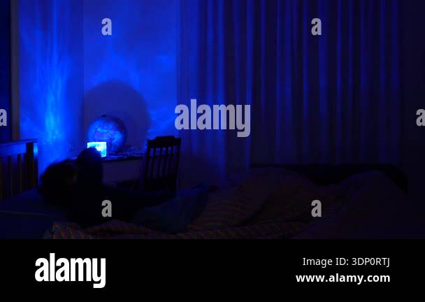 Teenage boy waking up in bed in dark room with blue lighting ...