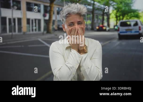 Woman with short hair covers mouth with hands and smiles shyly on city ...