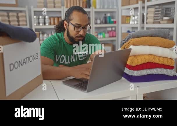 Man in volunteer shirt types on a laptop beside a donations box and ...