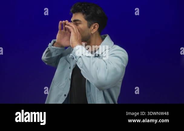 Young hispanic man cupping hands to mouth in studio with rich deep blue ...
